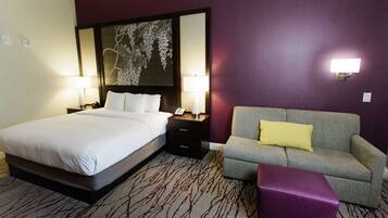 Quarto de hotel com uma cama grande, um sofá cinzento, um banquinho roxo pequeno e duas luminárias de cabeceira.