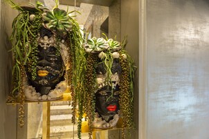 Duas máscaras decorativas ornamentadas, em preto e branco, com plantas verdes e elementos pendurados, exibidas sobre uma superfície de mármore.