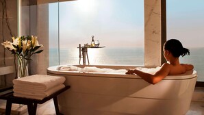 Mulher relaxando em uma banheira com vista para o mar. Há uma mesa com um vaso de flores e um tabuleiro com artigos de higiene pessoal.