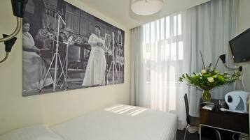 Quarto de hotel com uma fotografia grande em preto e branco na parede, uma cama, uma escrivaninha com um vaso de flores e uma televisão.