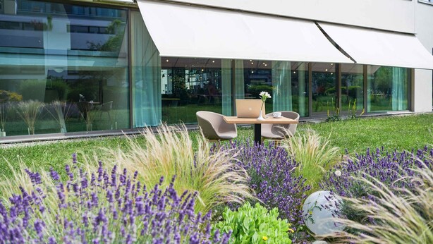 Um edifício moderno com grandes janelas, área coberta para sentar ao ar livre e um jardim bem cuidado com lavanda e outras plantas.