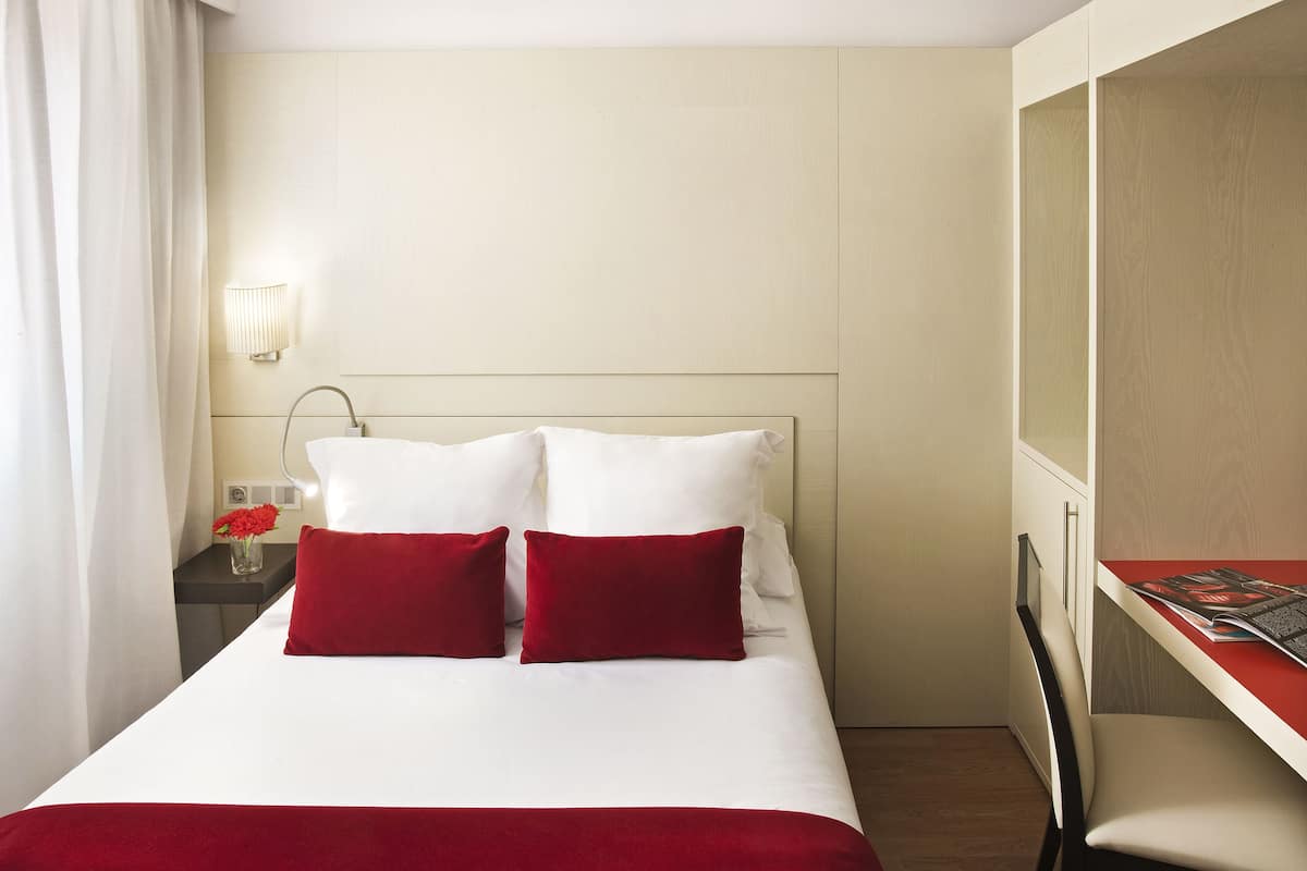 Quarto de hotel com cama, mesa de cabeceira, luminária, cadeira e escrivaninha.