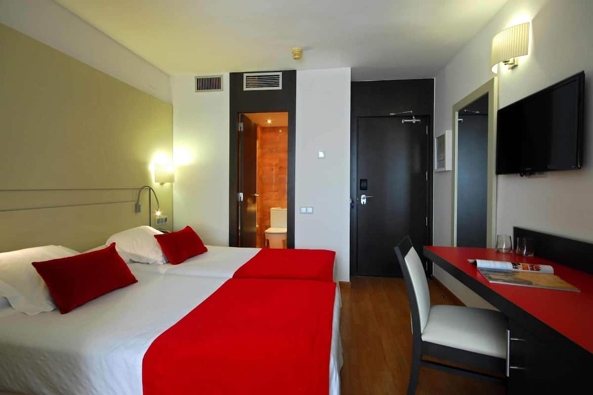 Quarto de hotel com duas camas, uma escrivaninha, uma cadeira, uma televisão e banheiro visível por uma porta aberta.
