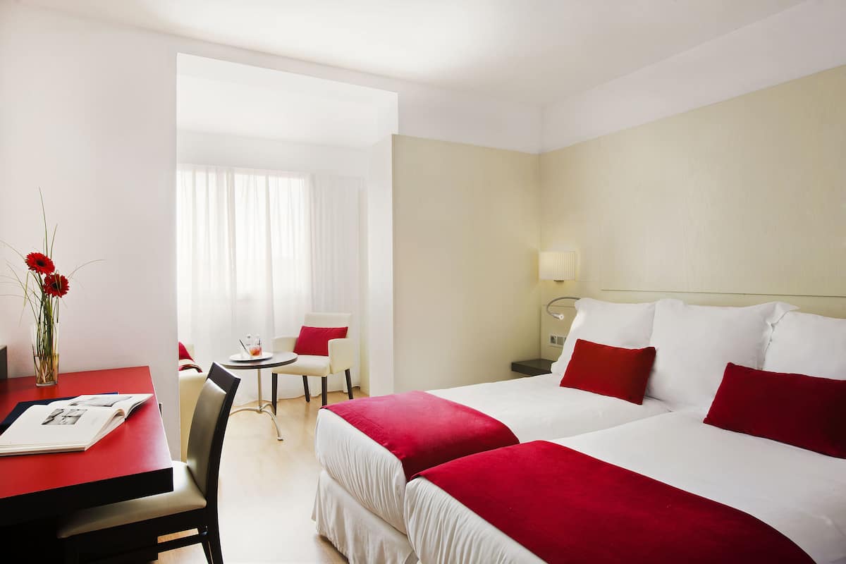Quarto de hotel com duas camas, uma mesa de jantar, uma cadeira e uma pequena mesa com abajur.