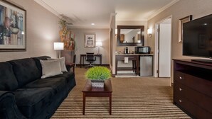 Quarto de hotel com sofá de cor escura, uma pequena mesa de madeira com uma planta, televisão, micro-ondas, frigorífico, secretária com cadeira e área de refeições com mesa e cadeiras.