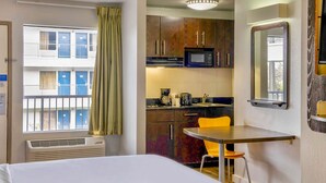 Quarto de hotel com cama, pequeno espaço de cozinha, mesa de jantar com cadeira, janela com cortinas e vista do edifício lá fora.