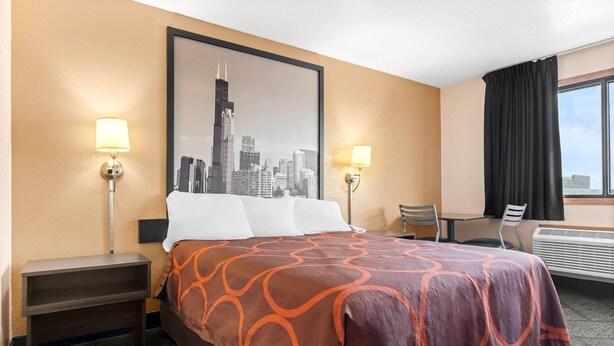 Quarto de hotel com cama, duas luminárias de cabeceira, uma pintura de paisagem urbana, uma mesa de jantar com cadeiras e uma janela com cortinas.