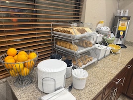 Um buffet de café da manhã com diversos pães, uma tigela de laranjas e um dispensador de massa para waffles.