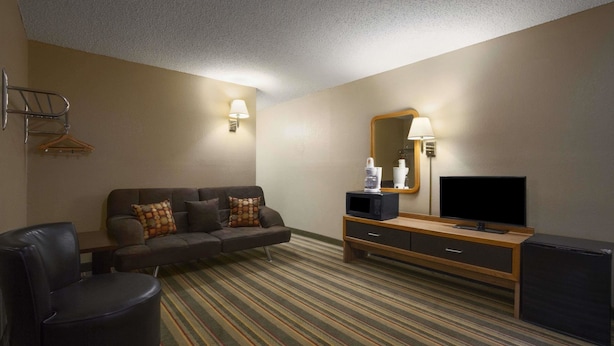 Quarto de hotel com sofá, cadeira, televisão, mini-geladeira, luminária e espelho.
