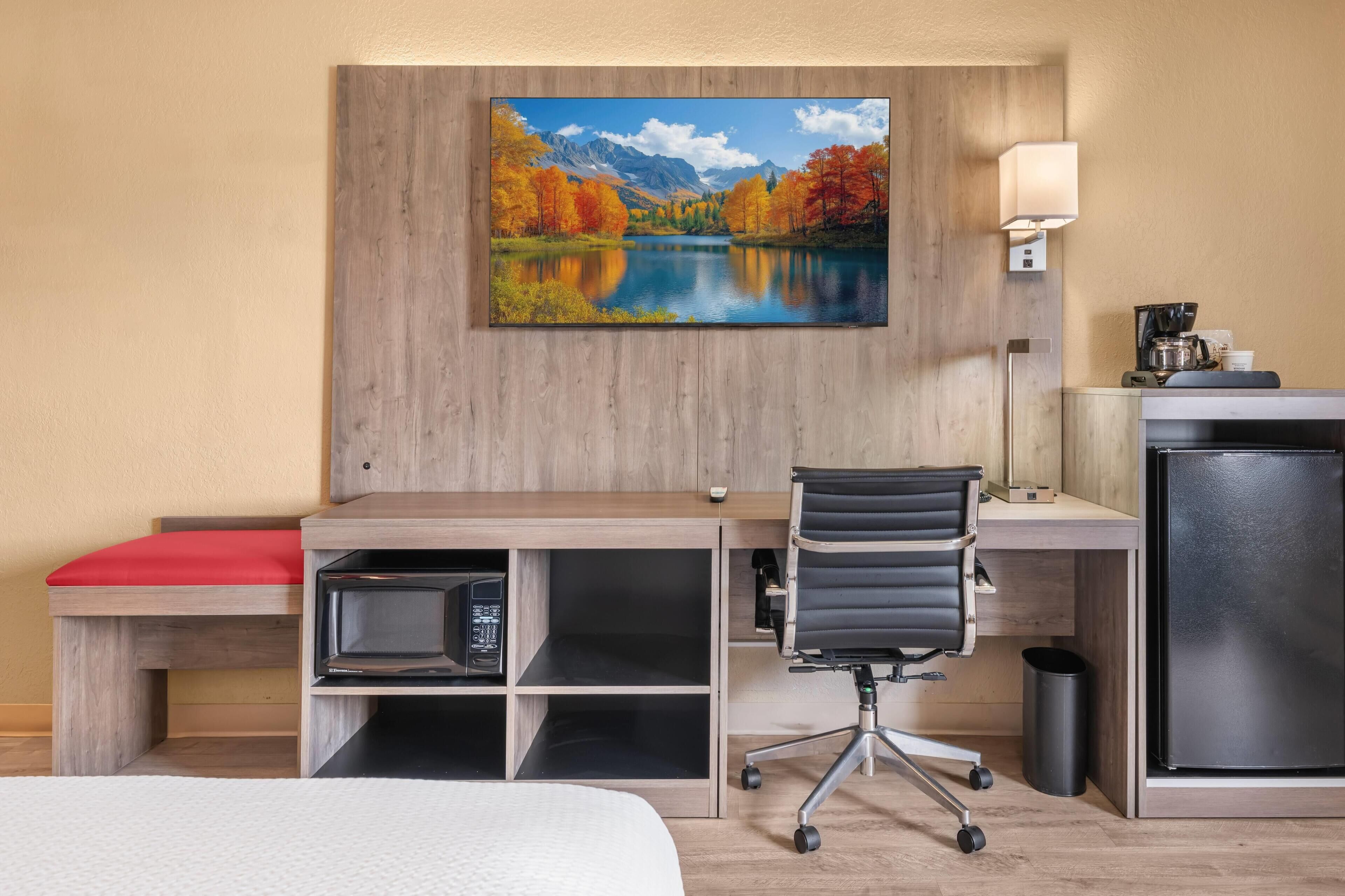 Quarto de hotel com escrivaninha de madeira, um banco vermelho, cadeira de escritório preta, micro-ondas, geladeira e pintura de um lago com árvores.