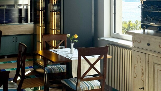 Uma área de refeições com uma mesa posta para dois, vista para o mar através de uma janela e uma cozinha com eletrodomésticos modernos.