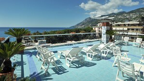 Piscina no terraço com espreguiçadeiras e vista para o mar e montanhas.