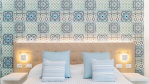 Cama bem arrumada com almofadas listradas, cabeceira com luzes fixadas na parede e parede revestida com azulejos estampados.
