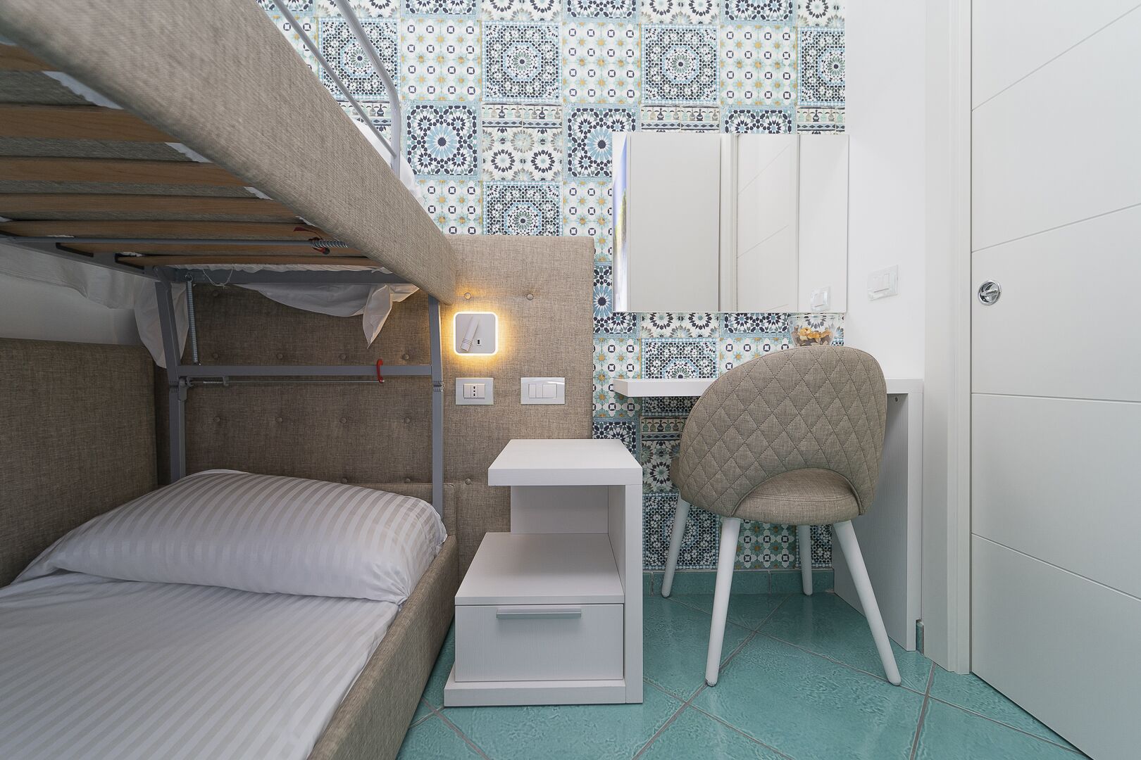 Quarto compacto com uma cama beliche, uma escrivaninha com cadeira, um pequeno armário branco e azulejos estampados nas paredes.