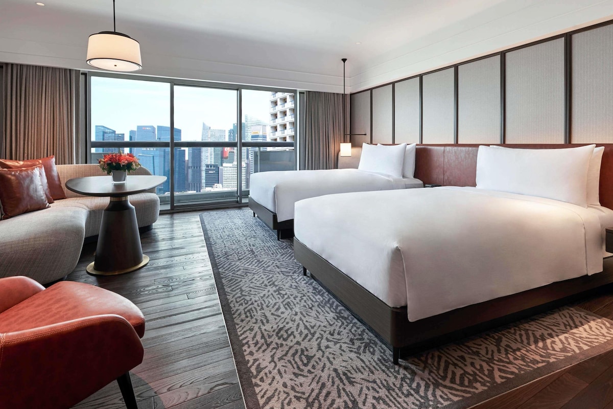 Quarto de hotel com duas camas, um sofá, uma mesinha de centro com um vaso e uma janela grande com vista para a cidade.