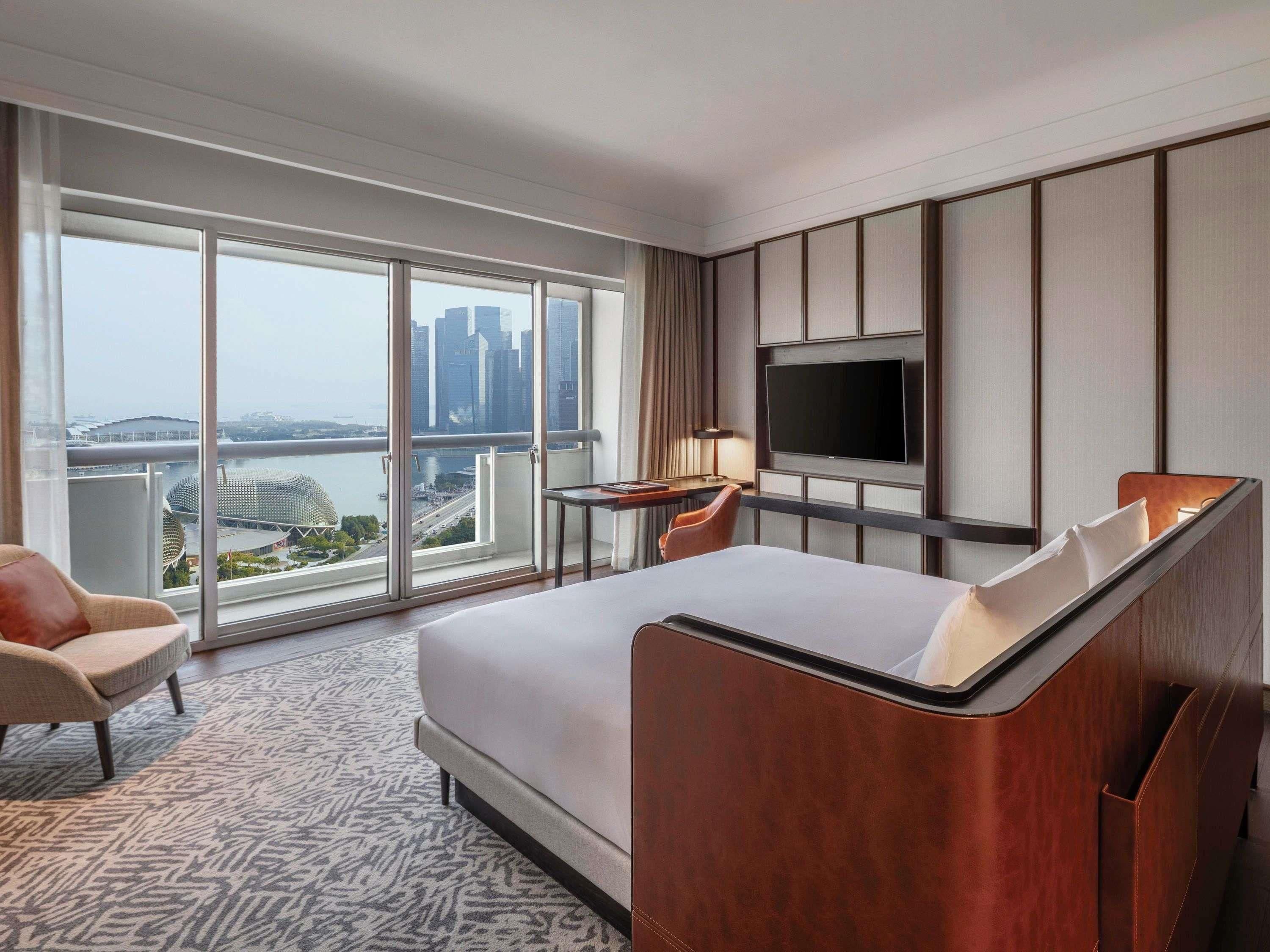 Quarto de hotel com uma cama grande, televisão, escrivaninha e cadeira. Oferece vista para a paisagem urbana através de amplas janelas.