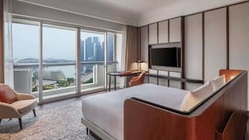 Quarto de hotel com uma cama grande, televisão, escrivaninha e cadeira. Oferece vista para a paisagem urbana através de amplas janelas.