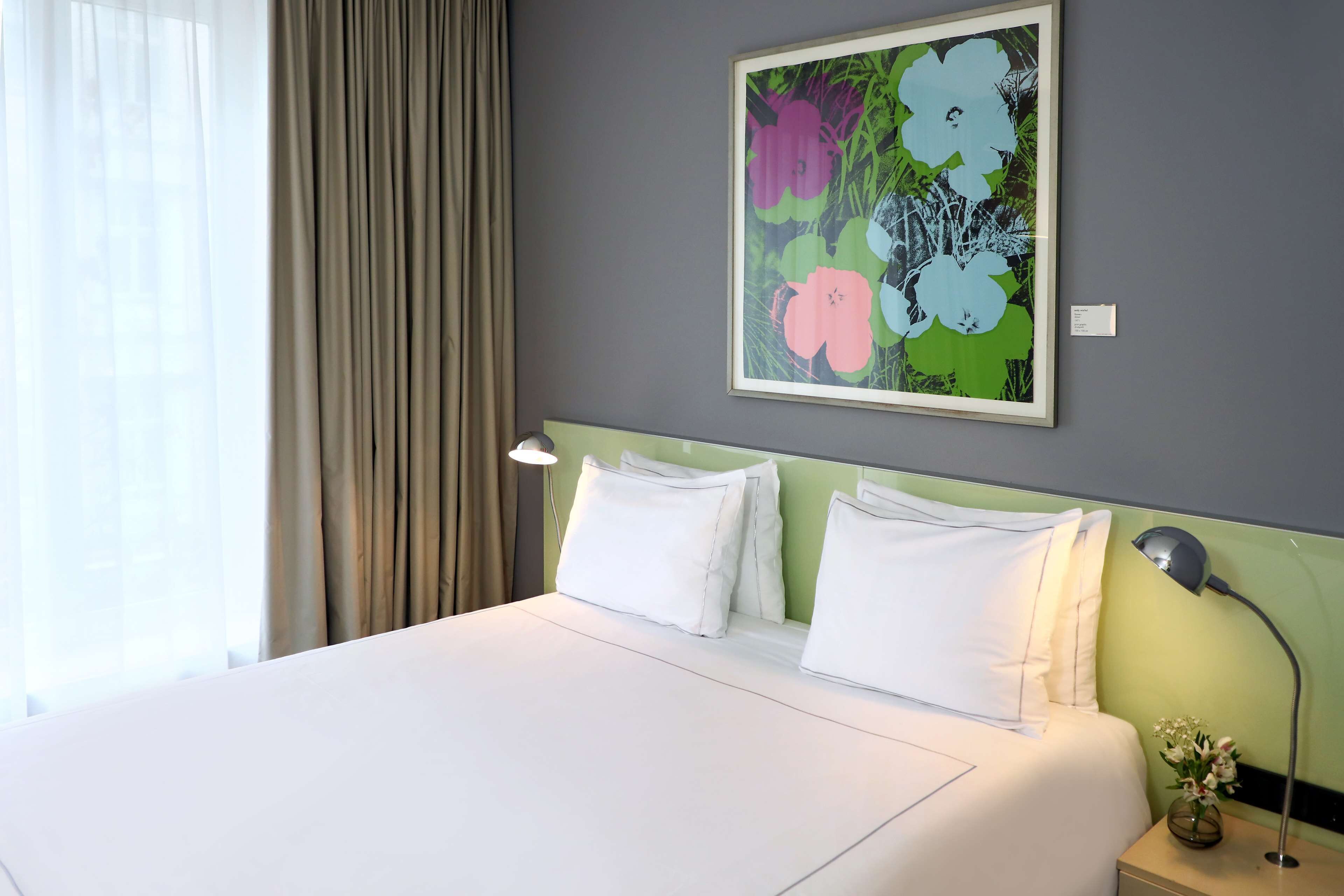 Quarto de hotel com uma cama grande, cabeceira verde, roupa de cama branca, uma obra de arte floral emoldurada na parede, um abajur e um vaso com flores na mesa de cabeceira.