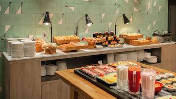 Buffet com diversos alimentos e bebidas, incluindo doces, frutas e outras opções, disposto em um balcão com parede de azulejos verdes ao fundo.