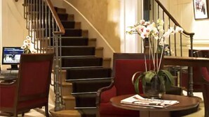 Um saguão de hotel com escadas, cadeiras estofadas em vermelho, uma mesa redonda com um vaso de flores e uma televisão em uma escrivaninha.