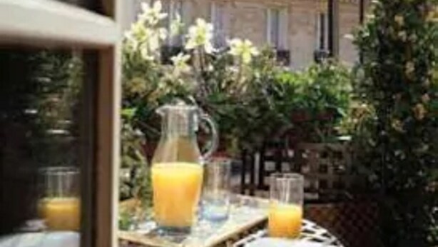 Varanda com mesa posta para dois, incluindo jarra e copos de suco de laranja, com vista para uma fileira de edifícios.