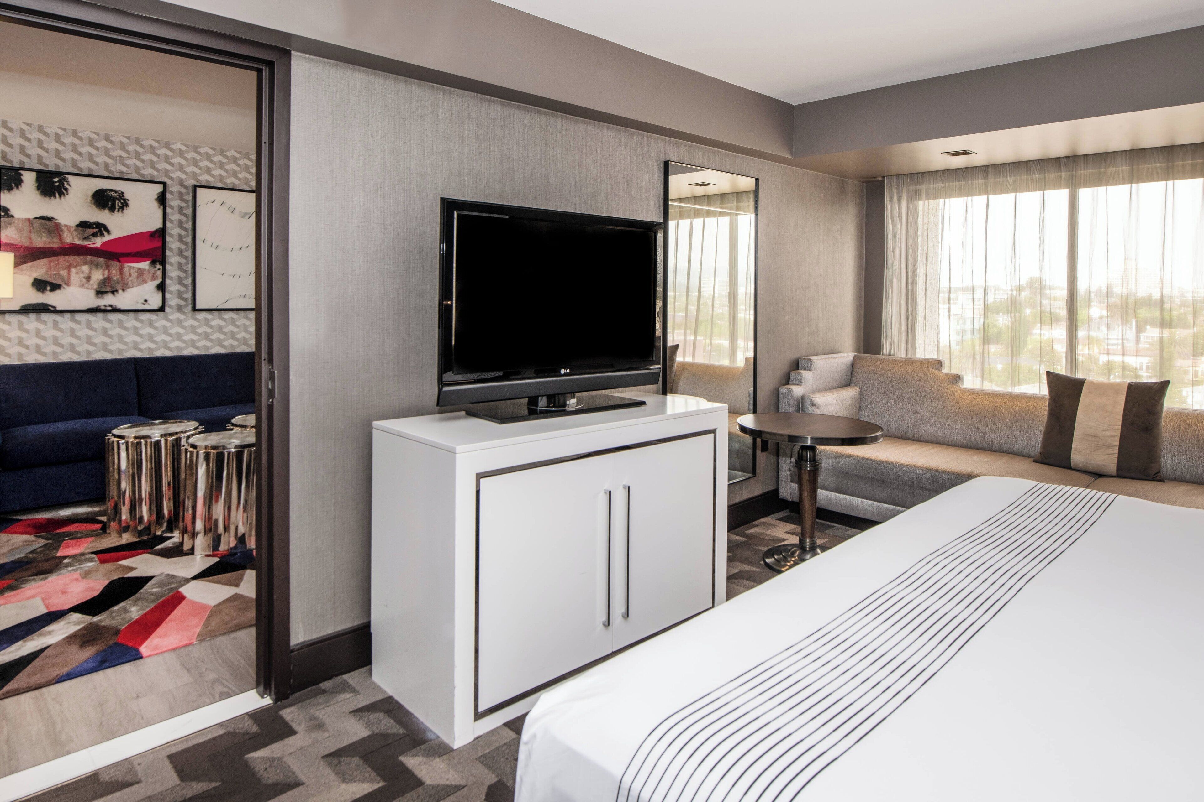 Quarto de hotel com cama, televisão sobre um móvel branco, sofá, mesa pequena e uma janela grande com cortinas.