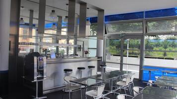 Interior moderno de restaurante com mesas e cadeiras altas, máquina de café e janelas amplas com vista para o exterior.