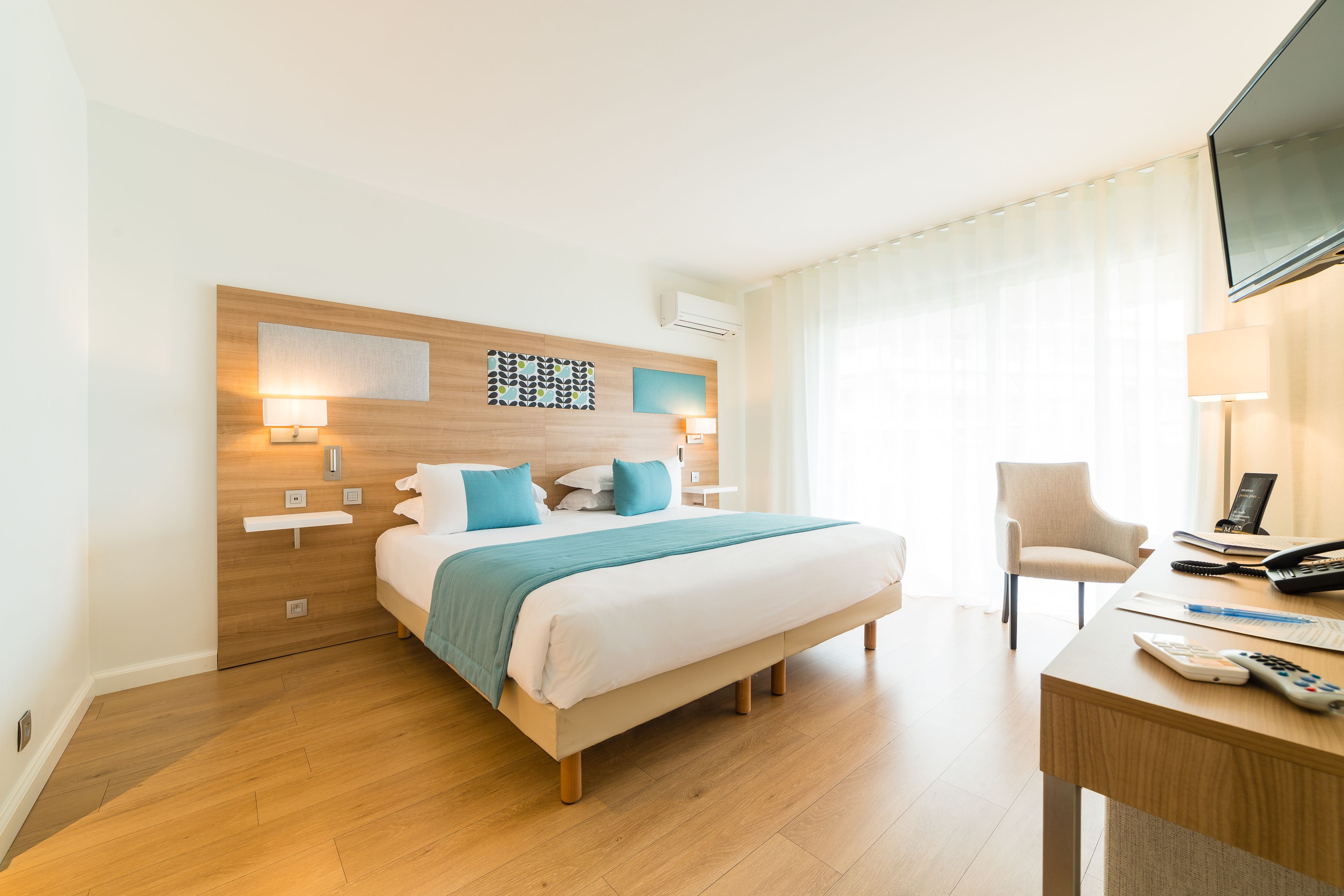 Quarto de hotel com cama, mesas de cabeceira, escrivaninha com cadeira, televisão e janela com cortinas.