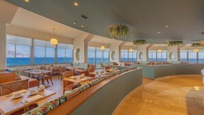 Um restaurante moderno com área de estar curvada, grandes janelas com vista para o oceano e plantas penduradas.