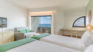 Quarto de hotel com duas camas, uma escrivaninha, uma cadeira, uma televisão e varanda com vista para o mar.