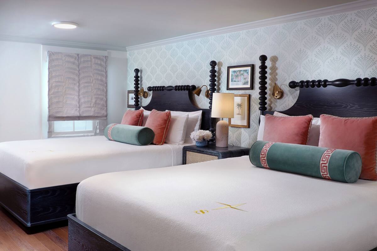 Quarto de hotel com duas camas, uma mesa de cabeceira com abajur, um quadro na parede e uma janela com cortina estampada.