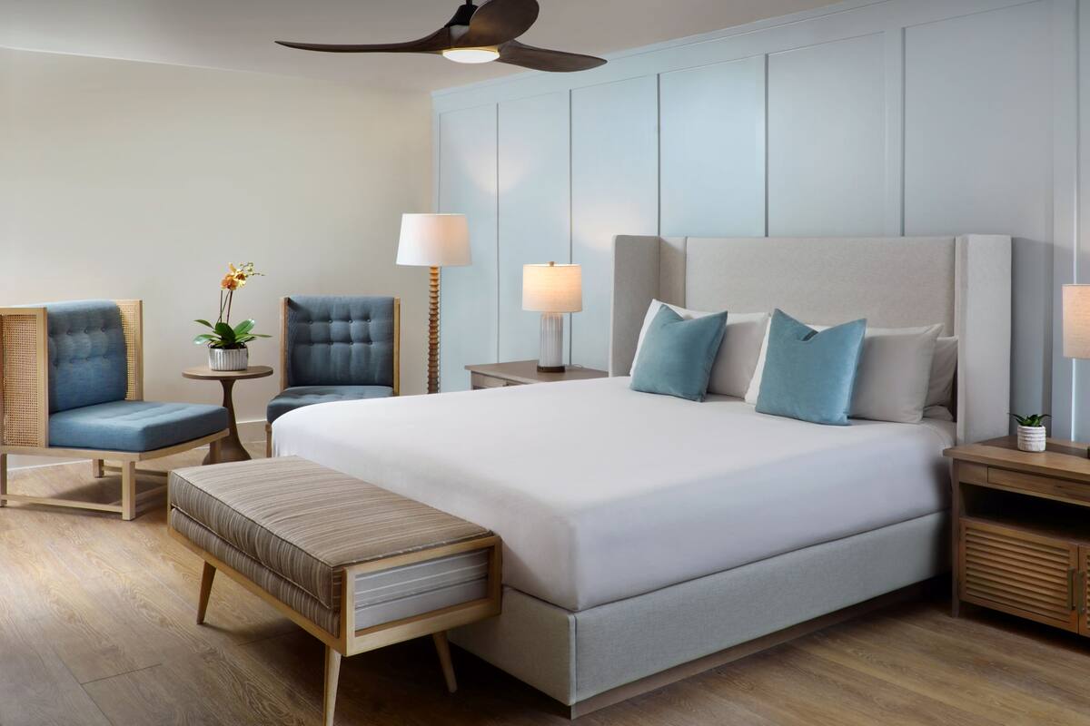 Quarto de hotel moderno com uma cama grande, duas mesinhas de cabeceira, uma cadeira, um banco e um ventilador de teto.