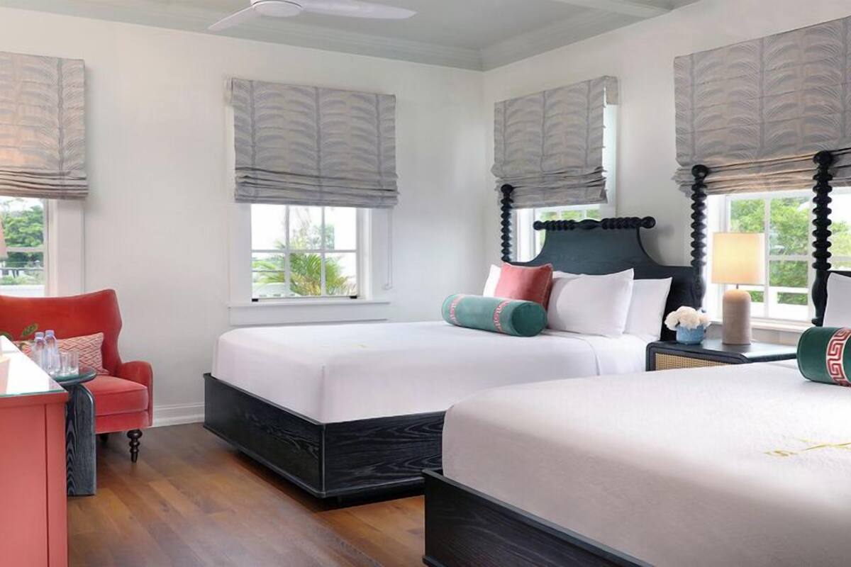 Quarto de hotel com duas camas, uma escrivaninha, uma cadeira e janelas amplas com cortinas.