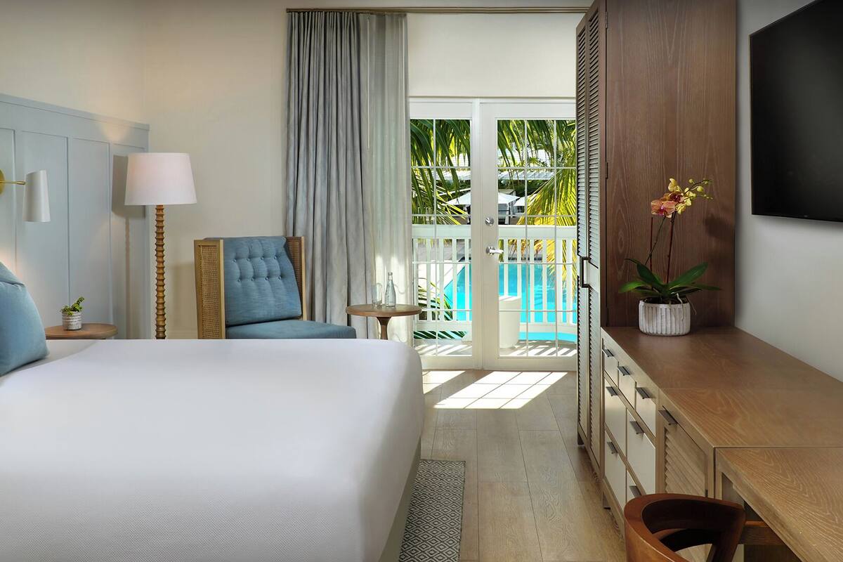 Quarto de hotel com cama, escrivaninha, cadeira, luminária, televisão e vista para uma piscina.