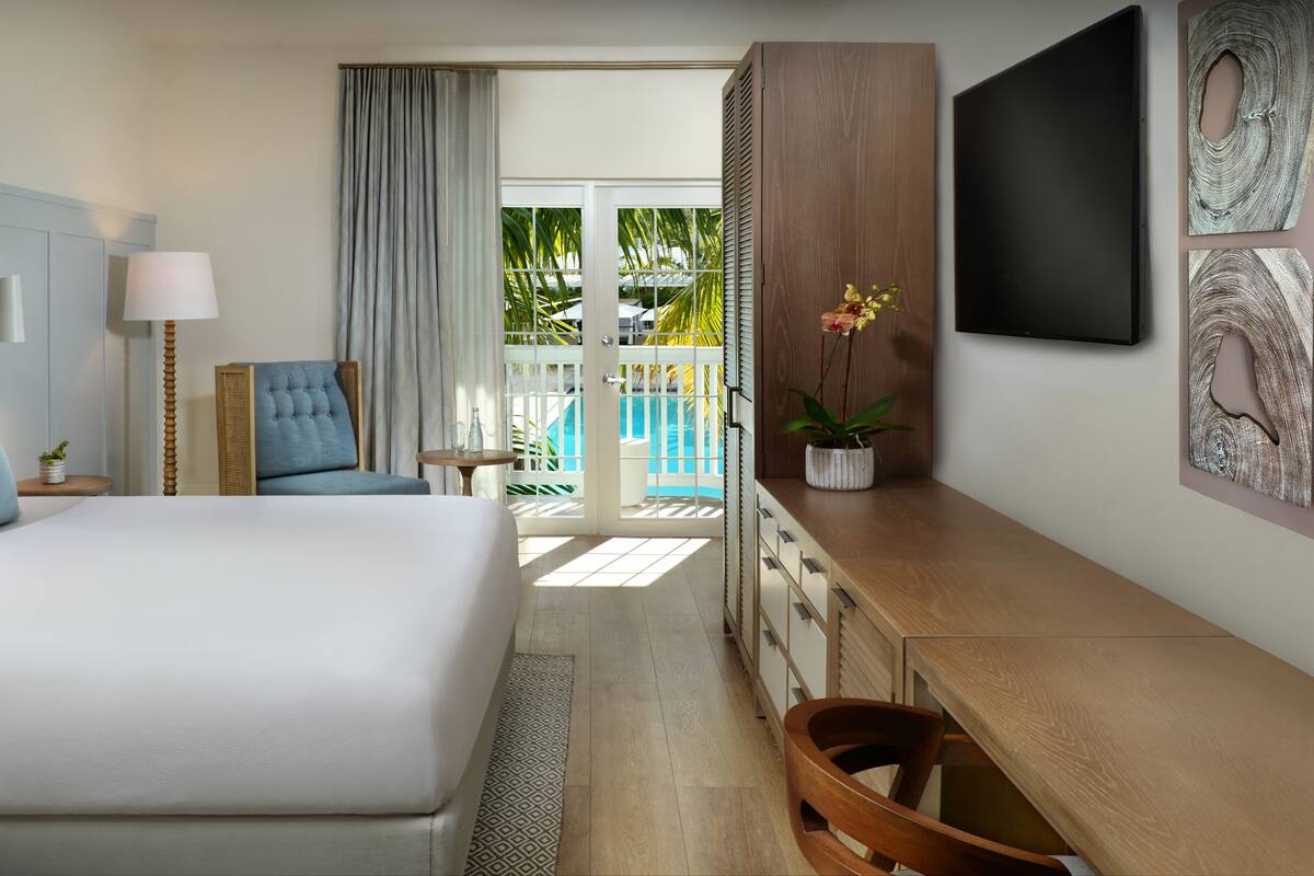 Quarto de hotel com uma cama grande, uma mesa, uma cadeira, uma TV e vista para uma piscina.