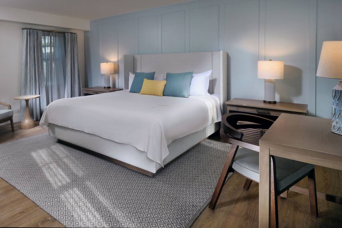 Quarto de hotel com uma cama grande, duas mesas de cabeceira com luminárias, uma cadeira, uma escrivaninha e um tapete estampado.