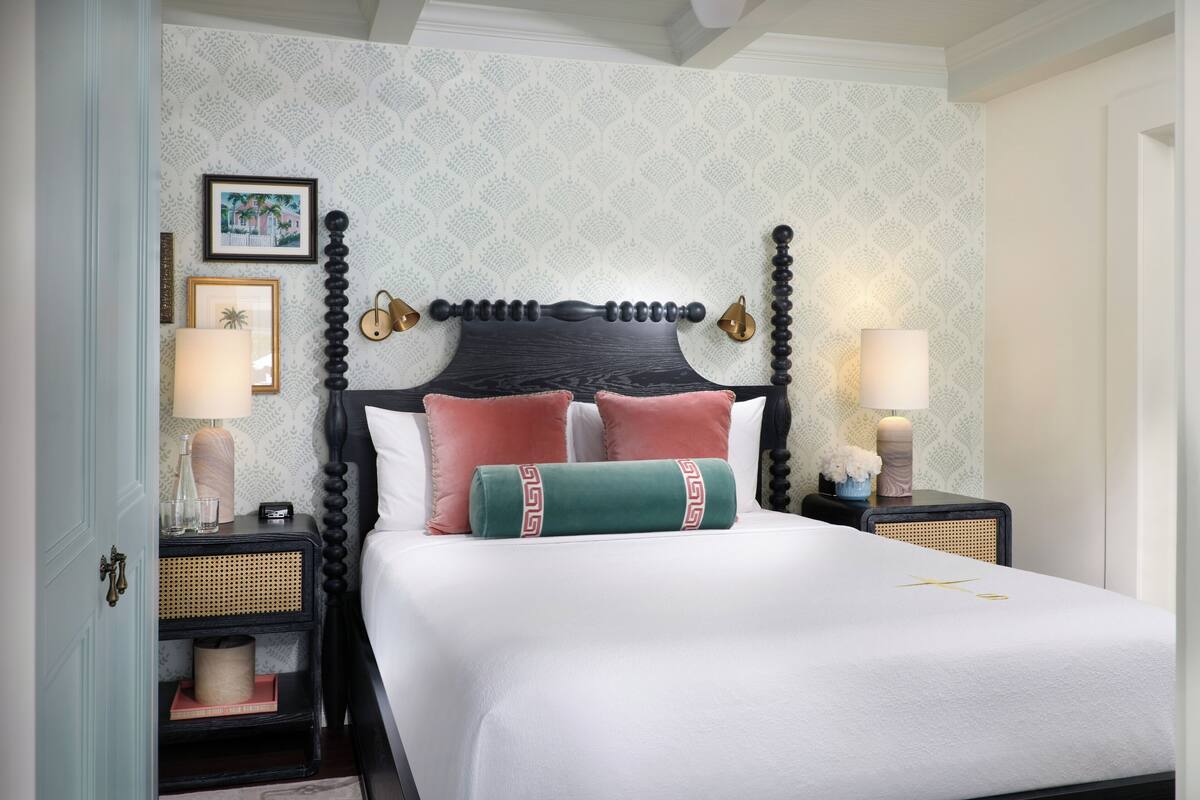 Cama bem arrumada com cabeceira preta, roupa de cama branca e almofadas decorativas. Há duas mesas de cabeceira com luminárias e um quadro na parede.