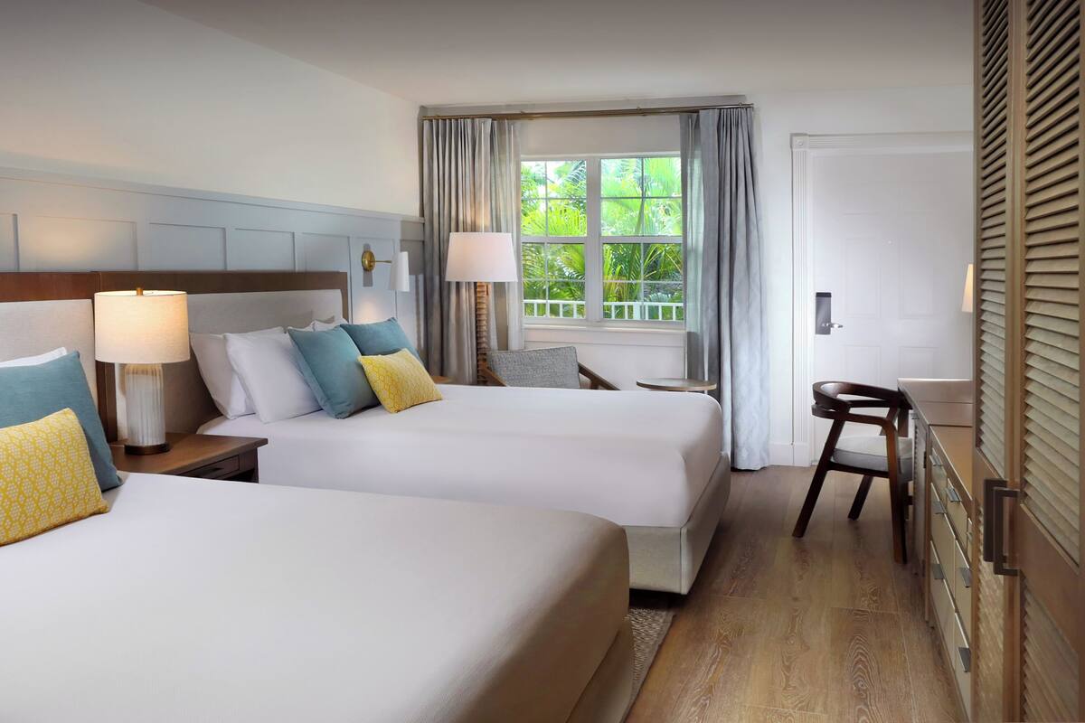 Quarto de hotel com duas camas, uma janela com cortinas, uma mesa de cabeceira com abajur e uma cadeira.