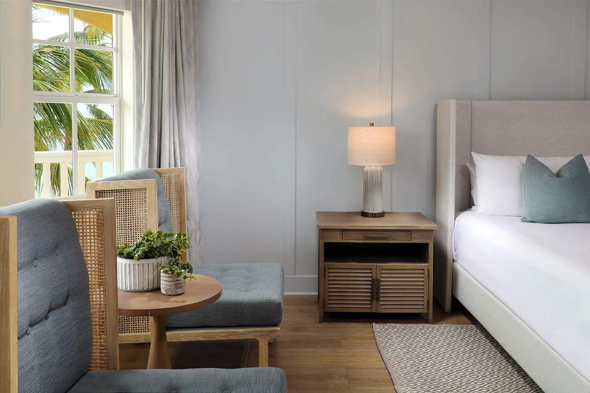 Quarto de hotel com uma cama, mesa de cabeceira com abajur, pequena mesa com planta, uma cadeira e uma janela com cortinas.