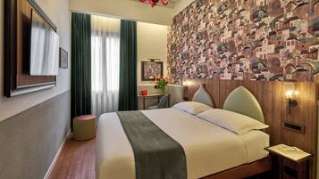 Quarto de hotel com uma cama grande, uma escrivaninha com cadeira, um banquinho pequeno, televisão presa à parede e parede com padrão.