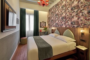 Quarto de hotel com uma cama grande, uma escrivaninha com cadeira, um banquinho pequeno, televisão presa à parede e parede com padrão.