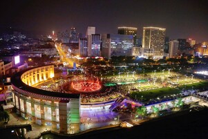 Um edifício grande e circular com uma fonte central, rodeado por uma praça com assentos e vegetação.