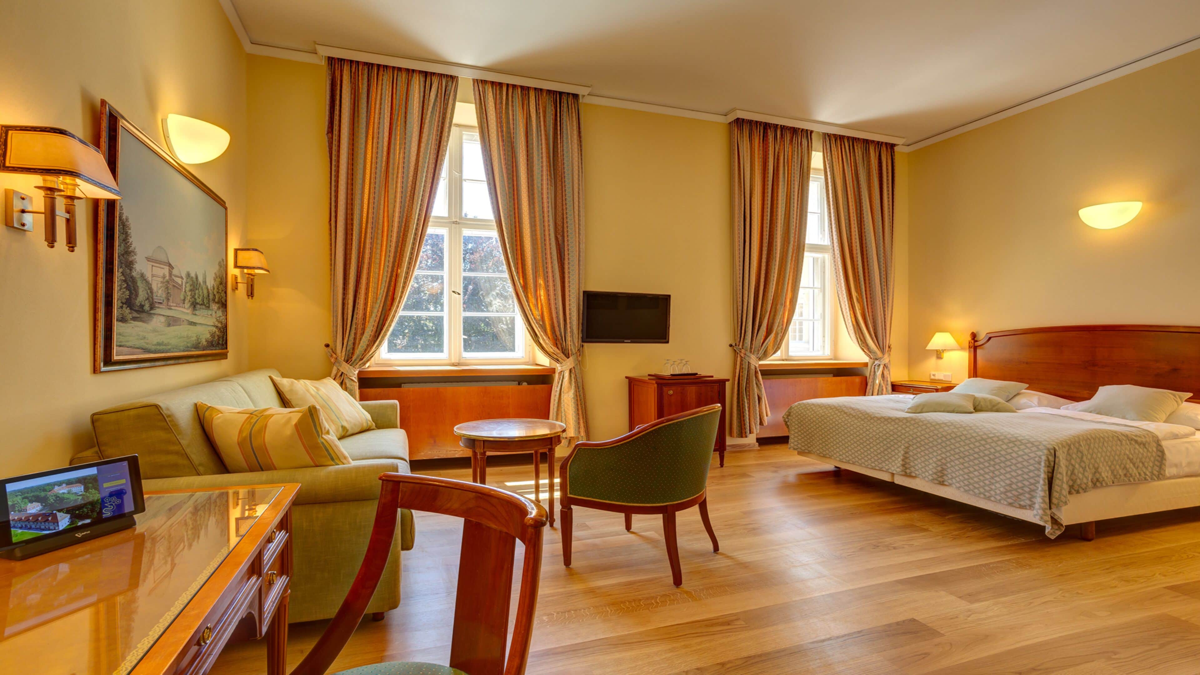 Quarto de hotel com cama, sofá, poltrona, mesa pequena e televisão.