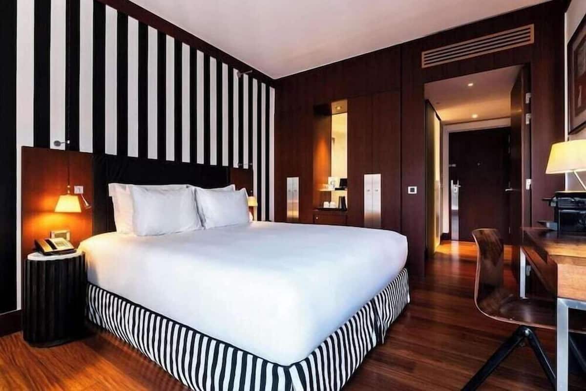 Quarto de hotel moderno com uma cama grande, mesa de cabeceira com abajur, uma cadeira e piso de madeira.