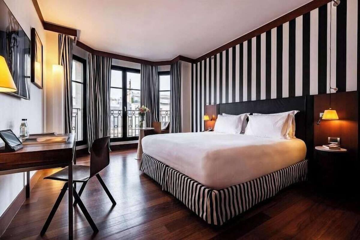 Quarto de hotel com uma cama grande, uma mesa de trabalho com cadeira, e vista para a cidade através de grandes janelas.