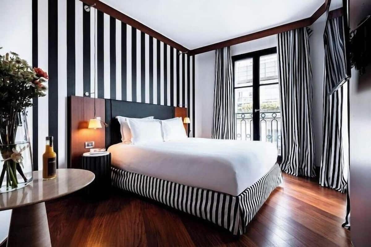 Quarto de hotel com uma cama grande, cortinas listradas, uma mesa redonda com um vaso e vista para o exterior.
