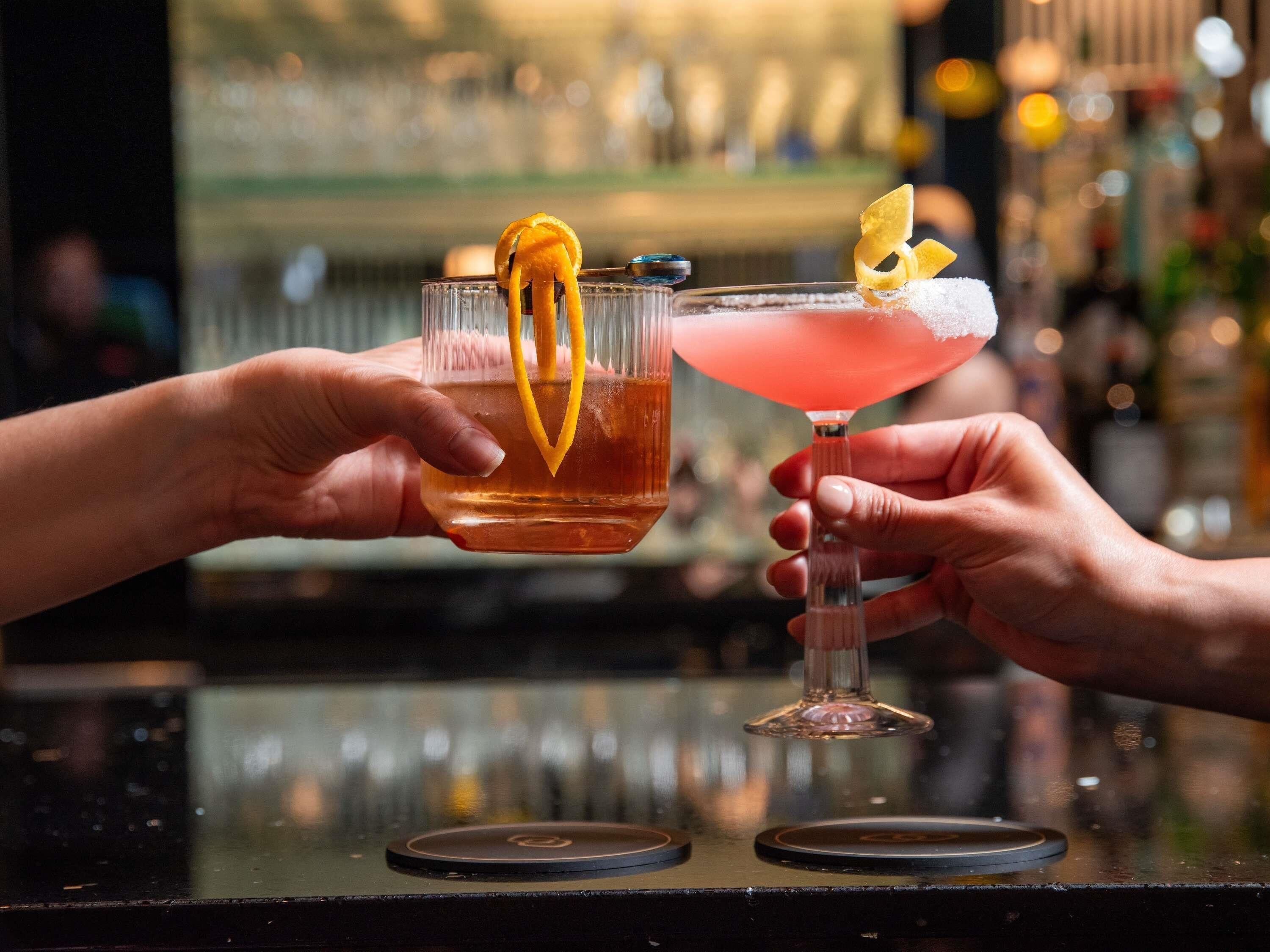 Duas mãos segurando copos de coquetel com guarnições, um balcão de bar e um fundo desfocado de um bar.