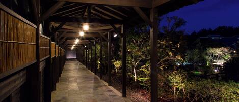 Uma galeria coberta com balaústres de madeira e pavimento de pedra, que conduz a um jardim com plantas e árvores iluminadas.