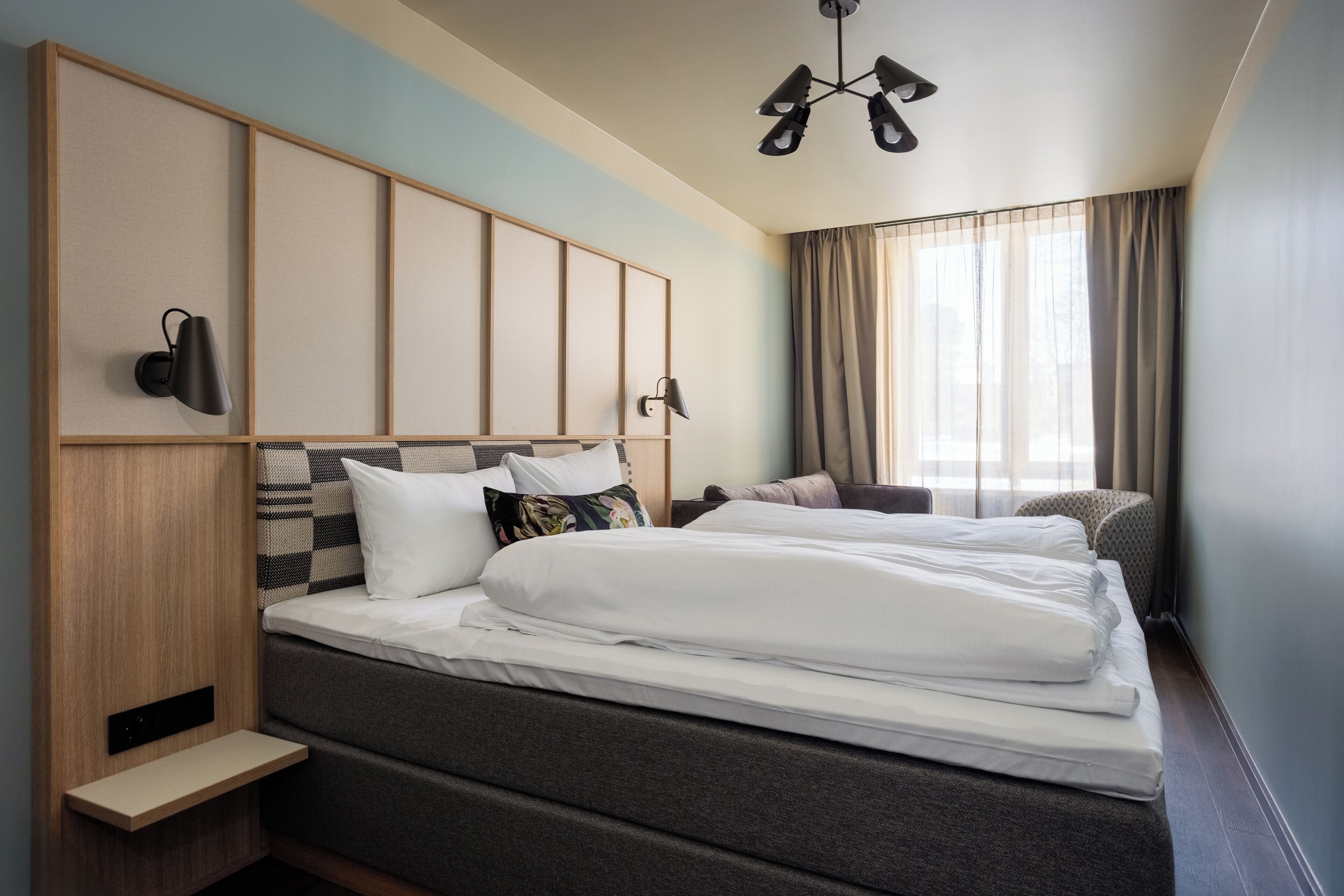 Quarto de hotel com uma cama grande, uma mesa de cabeceira, uma luminária na parede e uma janela com cortinas.