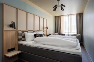 Quarto de hotel com uma cama grande, uma mesa de cabeceira, uma luminária na parede e uma janela com cortinas.